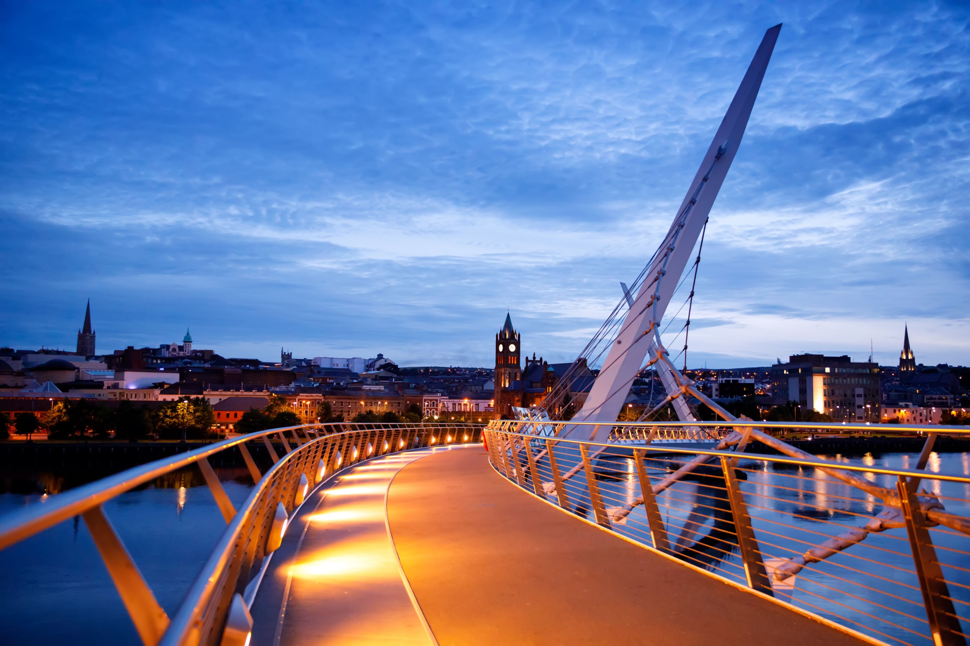 Uma ponte moderna com design em cabos atirantados, com vista para um horizonte urbano com imponentes torres de relógio e edifícios históricos.