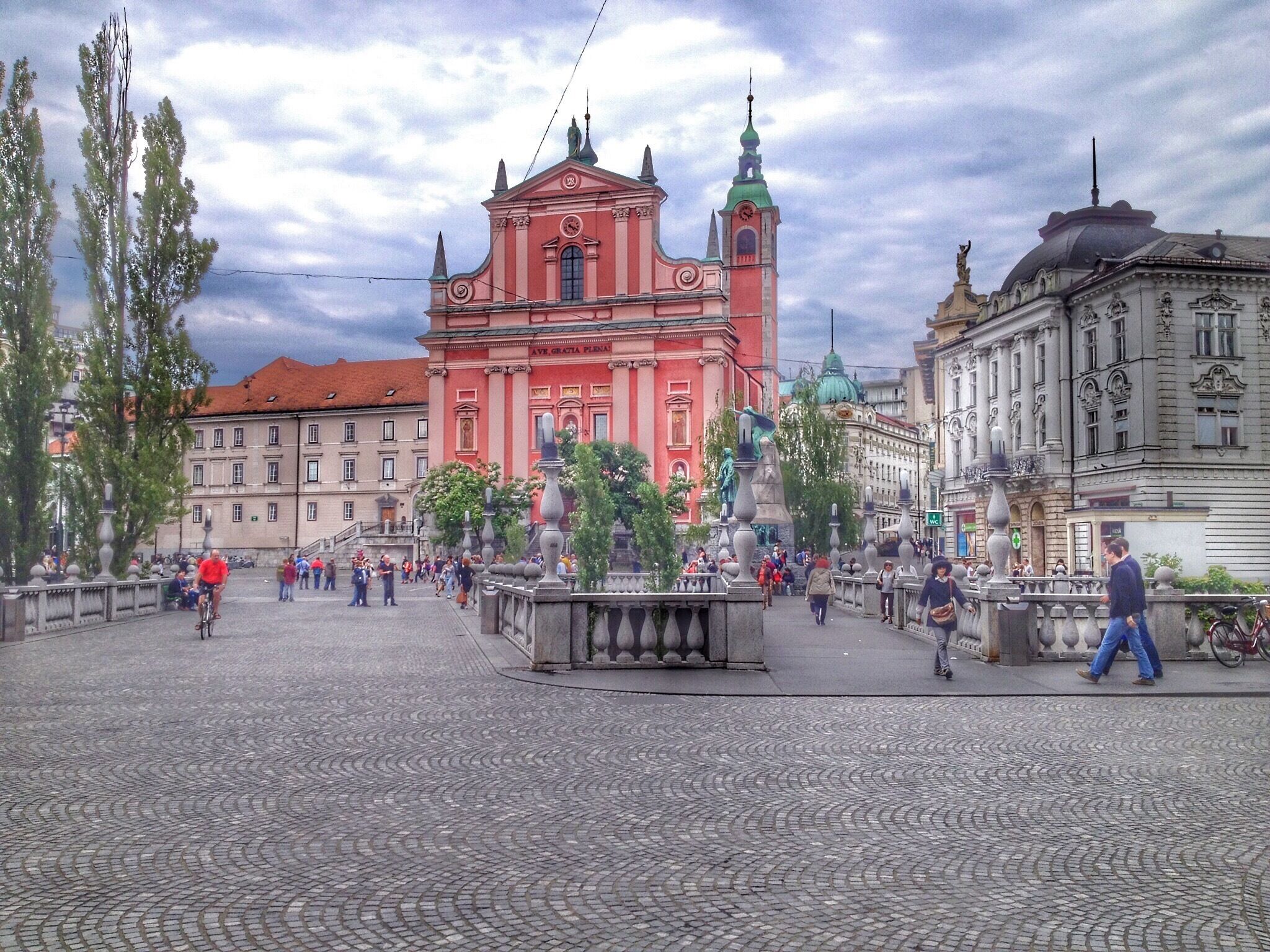 Uma praça de paralelepípedos com uma igreja rosa imponente, uma estátua e edifícios históricos ao redor.