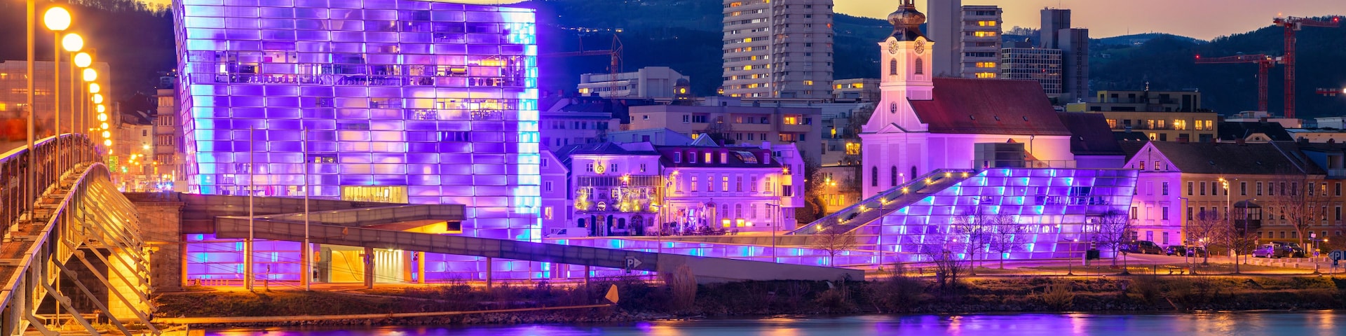 Paisagem urbana ao entardecer, com prédios iluminados, uma ponte e um rio refletindo as luzes.