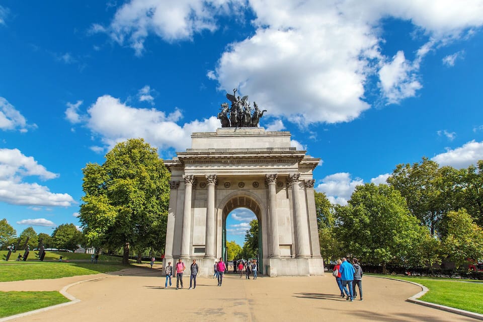 Um grande monumento em pedra com arco e estátua no topo, rodeado por árvores verdes e um céu azul límpido.