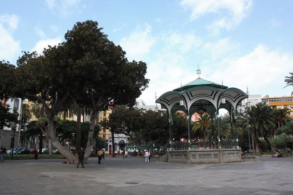 Uma praça com um coreto central, palmeiras e edifícios ao fundo.