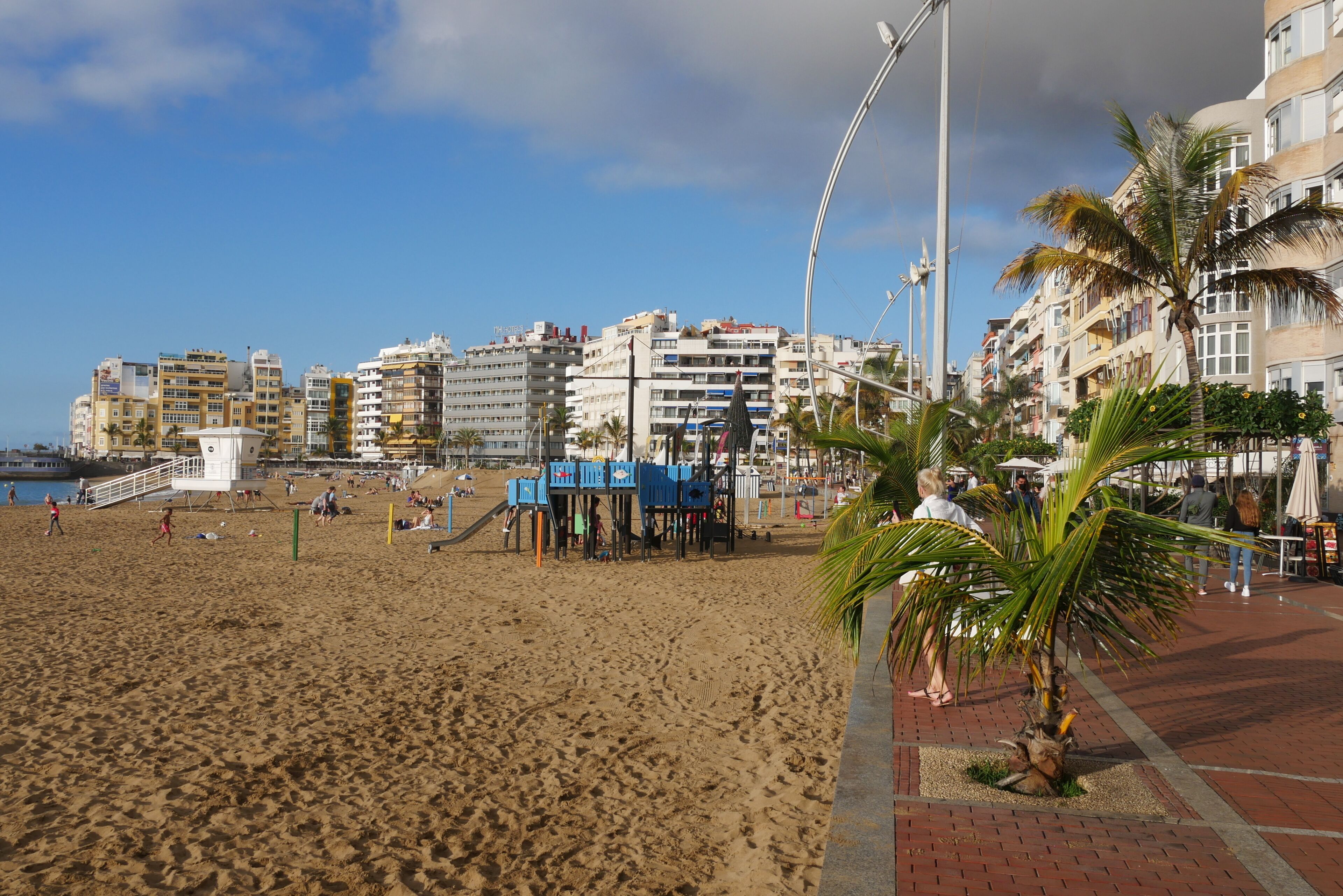 Uma praia arenosa com palmeiras, uma torre de salva-vidas e edifícios altos ao fundo.