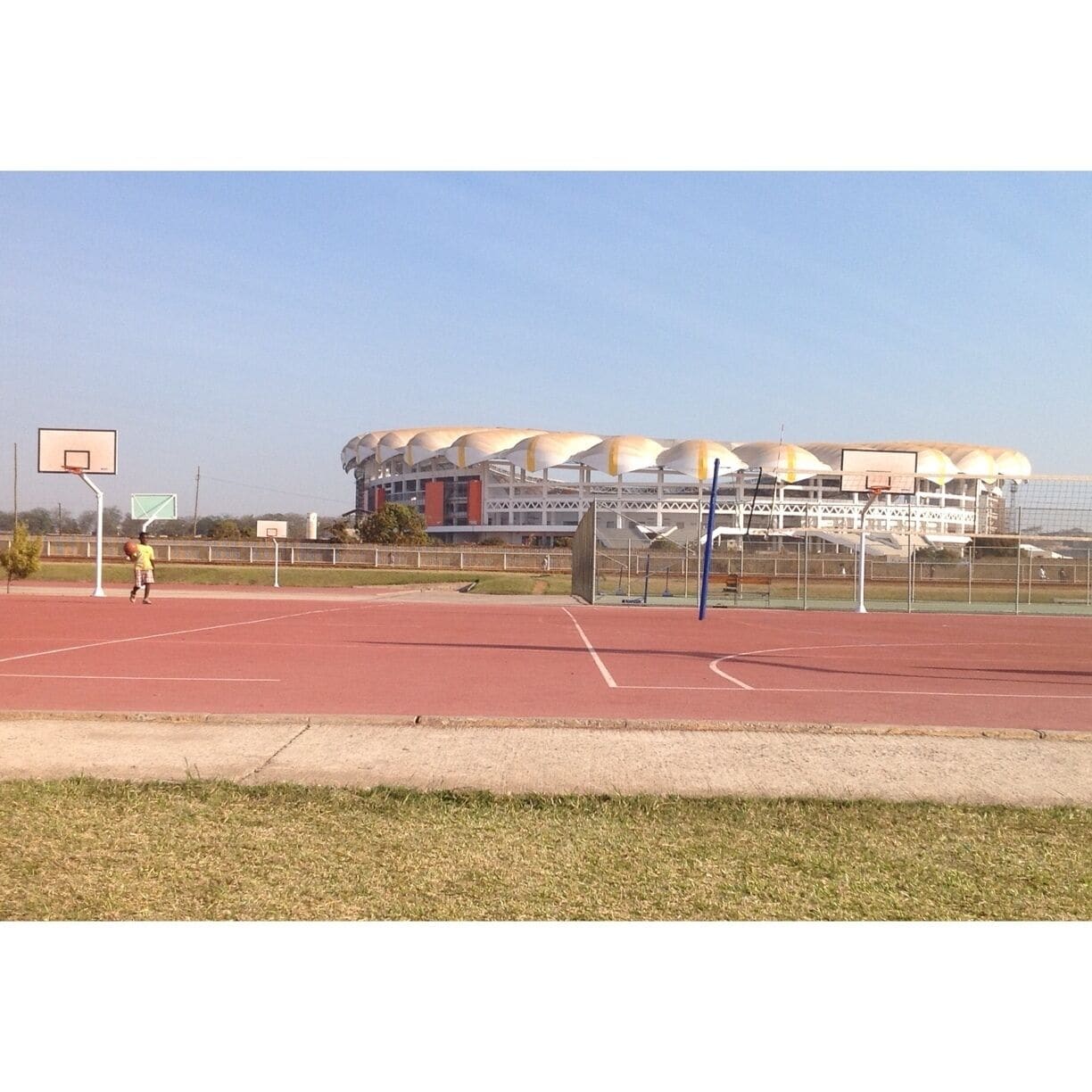 Uma quadra de basquete com uma pessoa jogando, uma instalação esportiva com um telhado característico e um céu claro.