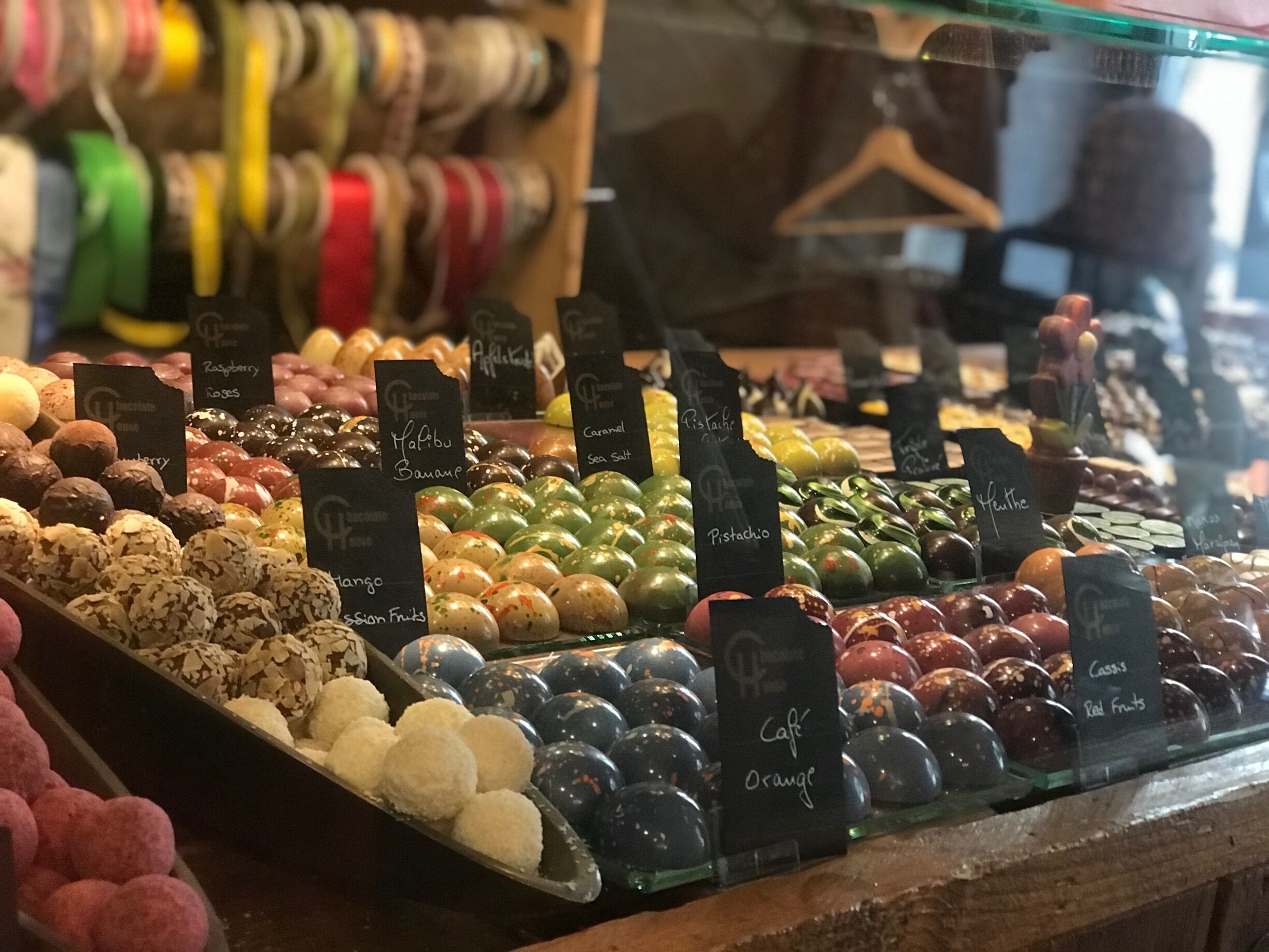 Diversos bombons coloridos expostos em vitrine de vidro, com etiquetas que identificam sabores como framboesa, manga e pistache.