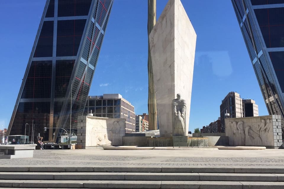 Uma praça moderna com um monumento central, rodeada por edifícios contemporâneos com fachada de vidro.