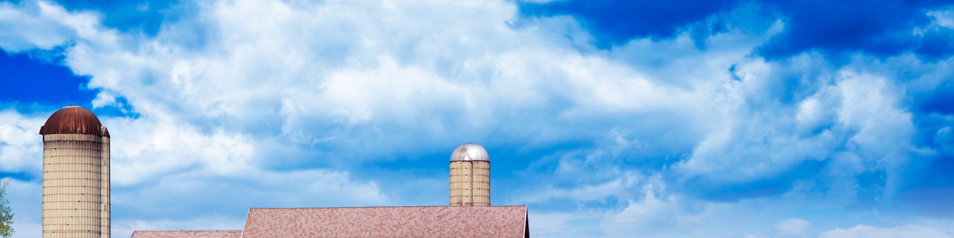 Um celeiro vermelho com um silo, cercado por campos verdes sob um céu azul com nuvens.