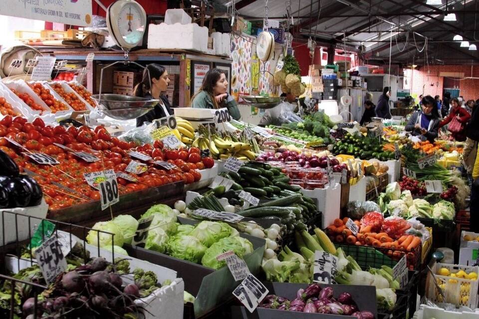 Um mercado interno movimentado com diversos produtos frescos à mostra, como tomates, pepinos e folhas verdes.