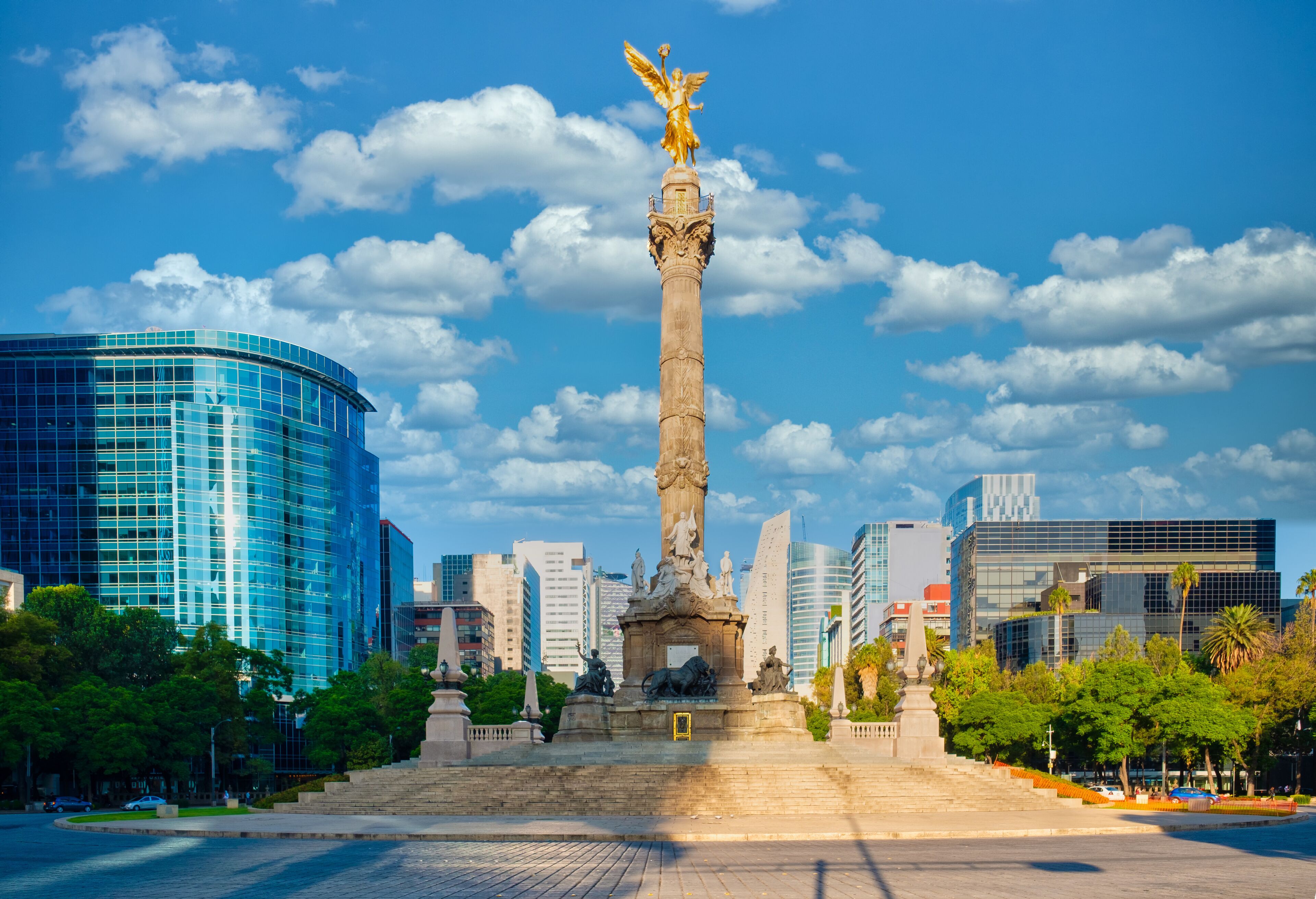 Um monumento alto com uma estátua dourada, rodeado por edifícios modernos e árvores.
