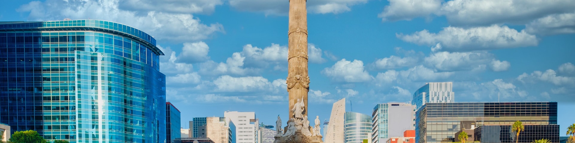 Um monumento alto com uma estátua dourada, rodeado por edifícios modernos e árvores.