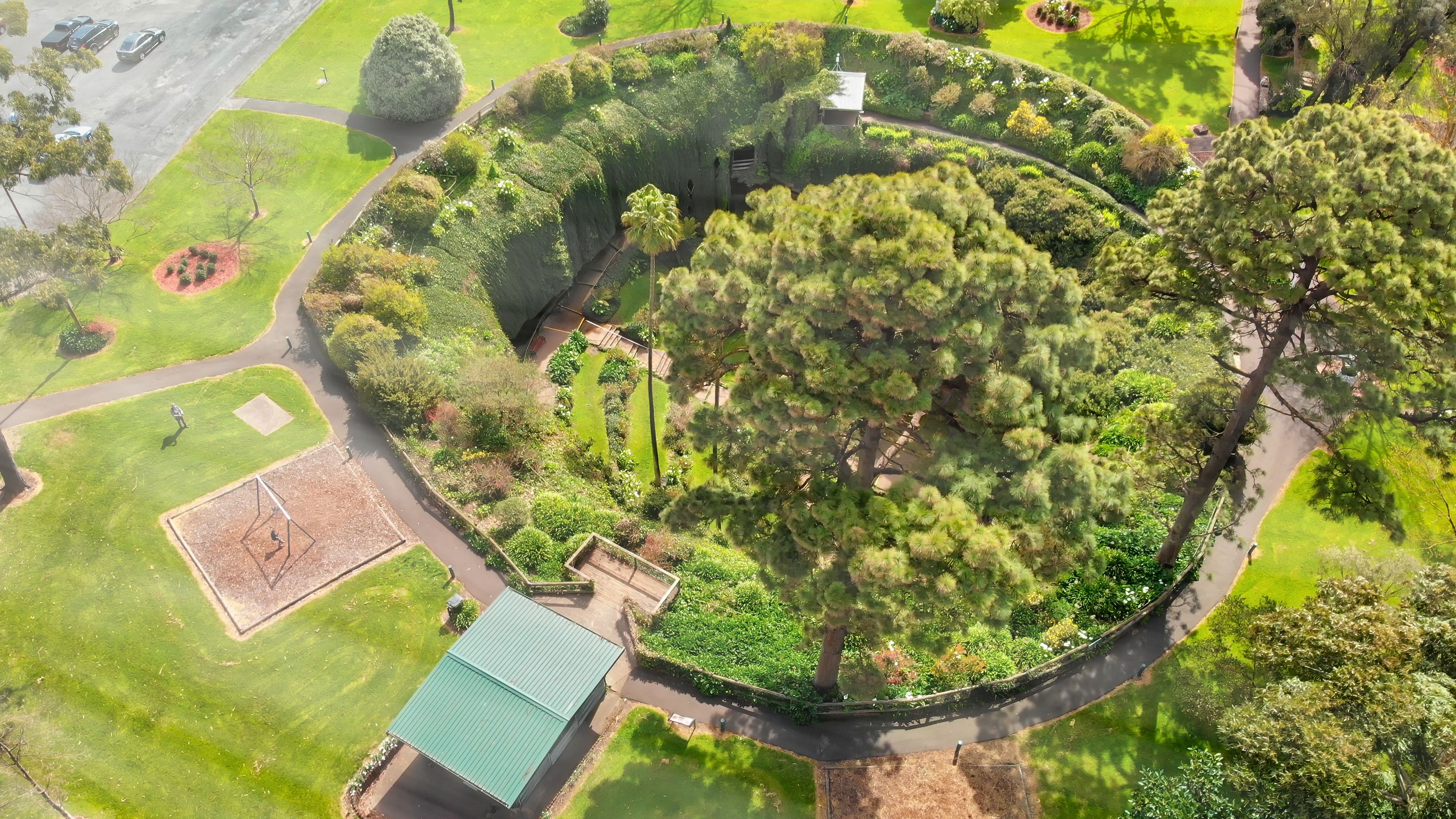 Vista aérea de um parque com disposição circular, uma árvore central e um pequeno edifício com telhado verde.