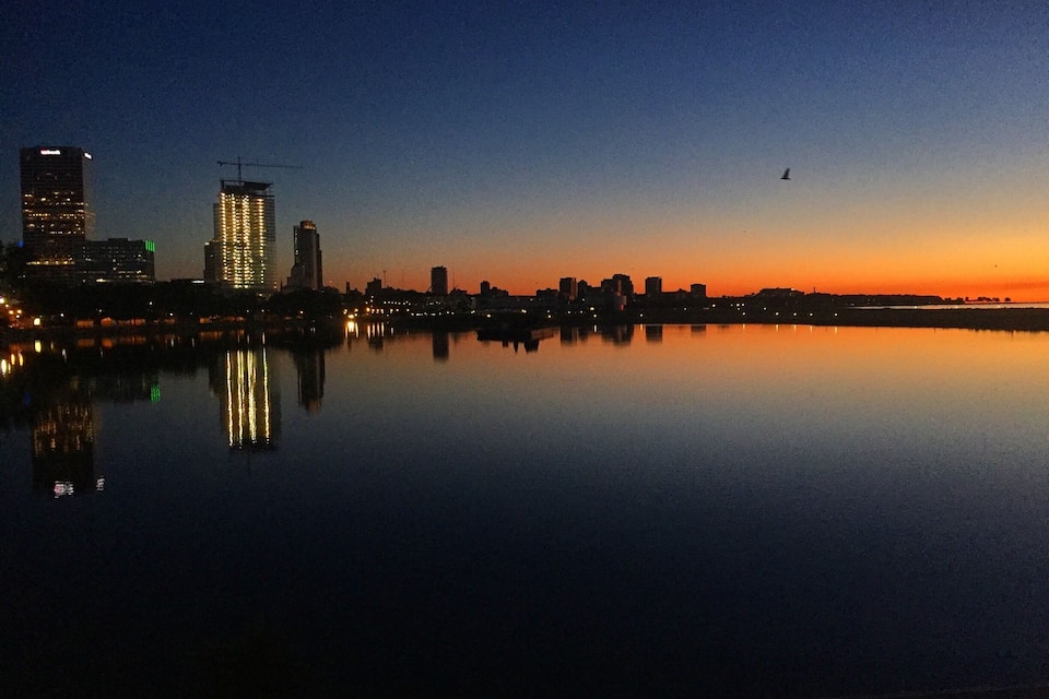 Silhueta urbana ao entardecer, com edifícios refletidos em águas calmas.