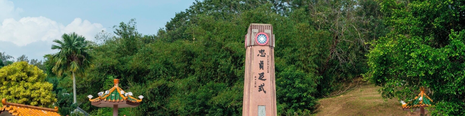 Um pavilhão tradicional chinês com telhado de telhas vermelhas, rodeado por um jardim com azulejos coloridos e um monumento de pedra com caracteres chineses.