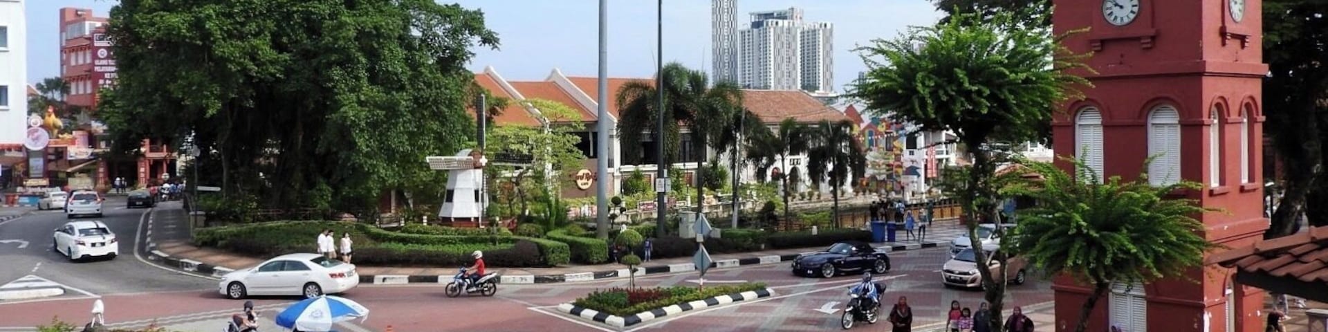 Um movimentado cruzamento urbano com uma rotatória, uma torre do relógio vermelha e um carro alegórico decorativo em primeiro plano.