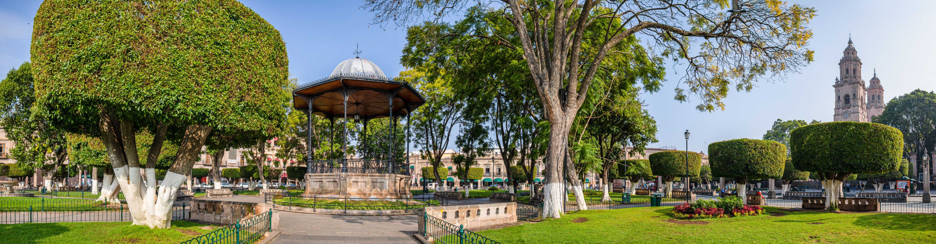Um parque com um coreto, árvores bem aparadas, uma fonte e um edifício histórico ao fundo.