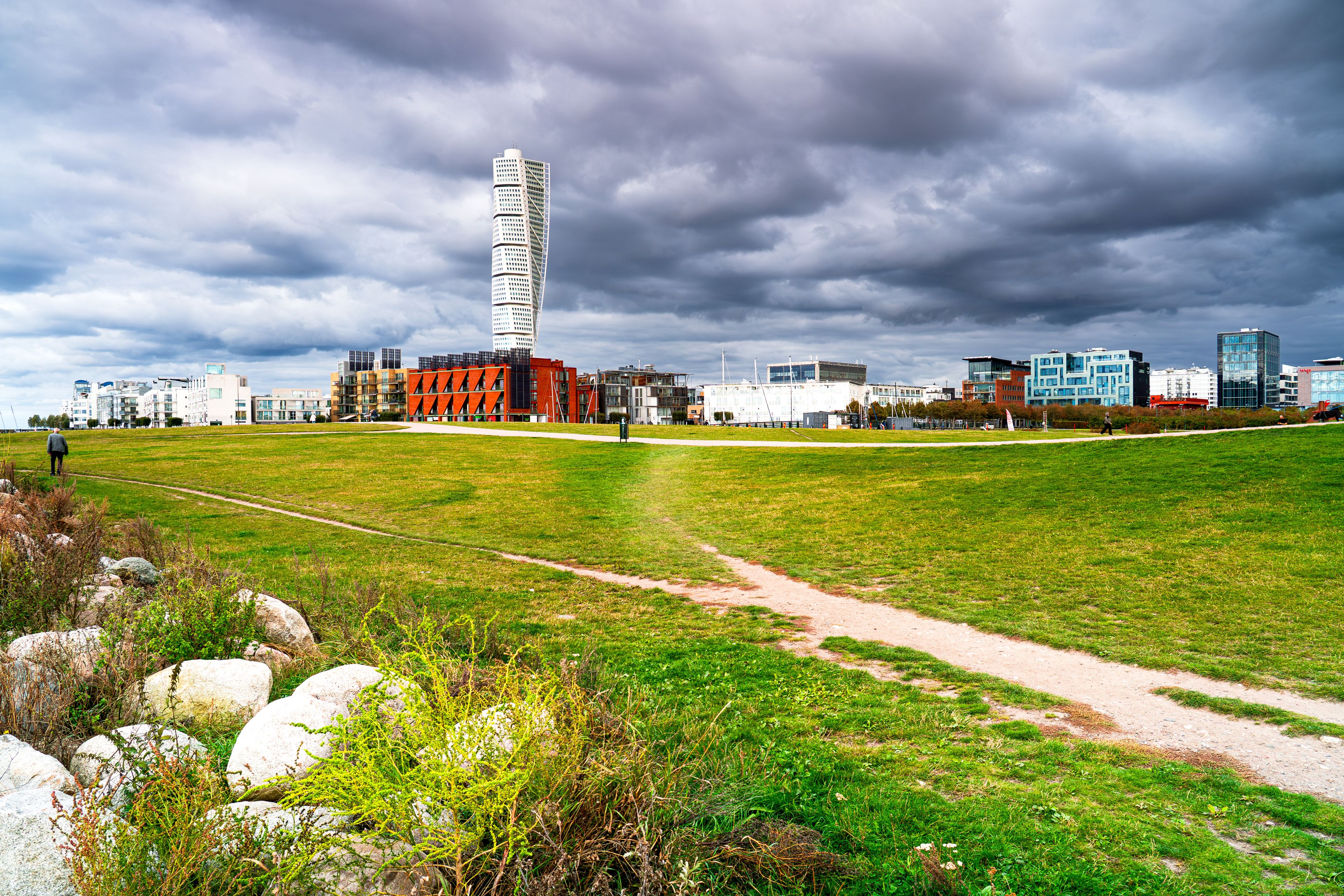 Um campo gramado com um caminho sinuoso, uma torre alta e edifícios modernos ao fundo.