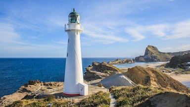 Um farol branco com cobertura verde, localizado em um trecho rochoso da costa com águas azul-claras.
