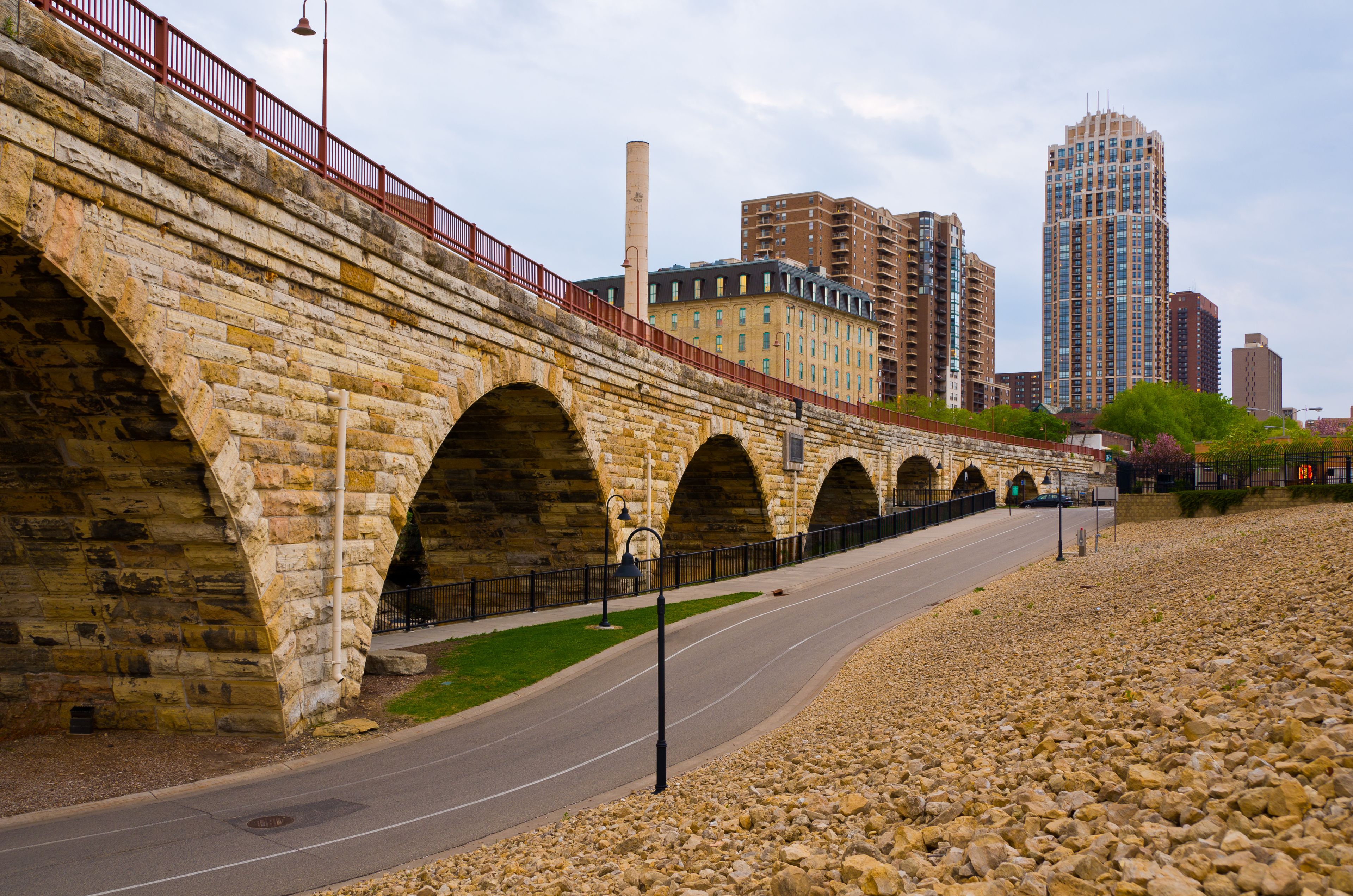 Uma ponte de pedra com várias arcos, uma estrada pavimentada e edifícios modernos ao fundo.