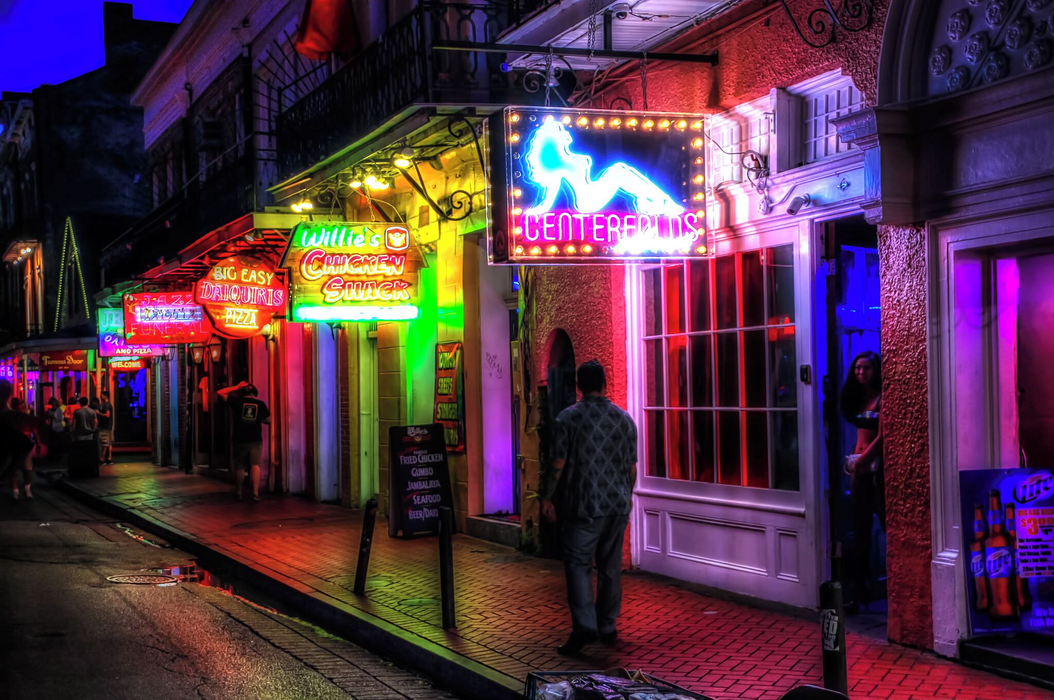 Uma rua bem iluminada com letreiros de neon para vários restaurantes, incluindo um de frango frito e outro de pizza.