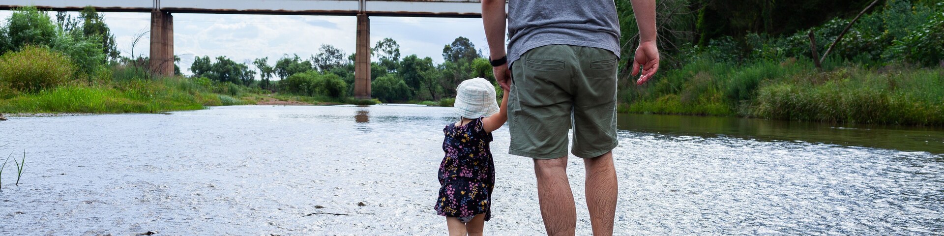 Um homem e uma criança caminham por um rio raso, com uma ponte ao fundo.