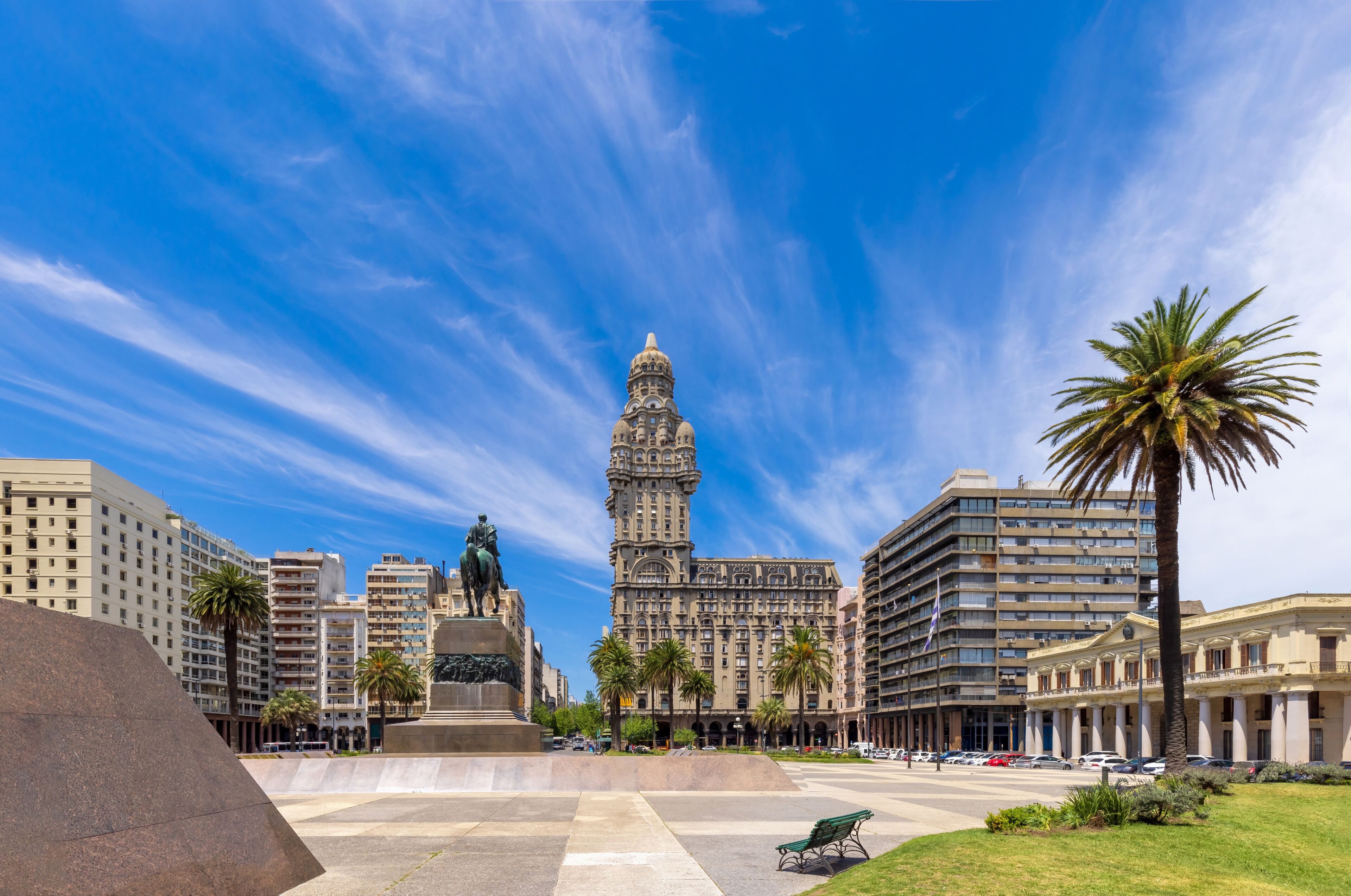 Uma praça com uma torre imponente, palmeiras e edifícios modernos.