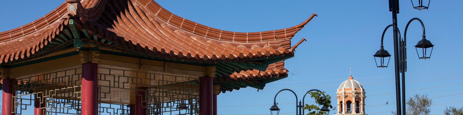 Um pavilhão tradicional com telhado curvo, pilares vermelhos e lanternas penduradas. Ao fundo, uma torre de sino e um céu azul límpido.