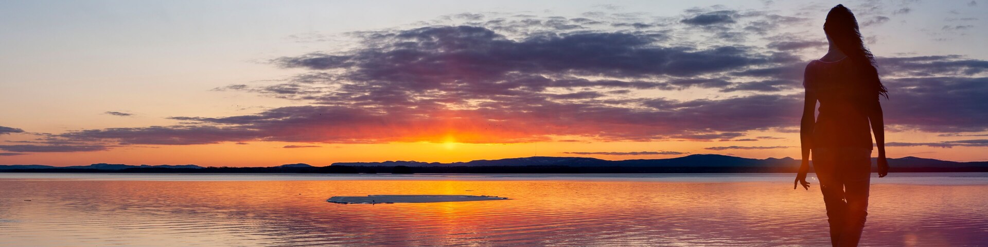 Uma pessoa em pé sobre uma superfície aquática refletora durante o pôr do sol, com o perfil de montanhas ao fundo.