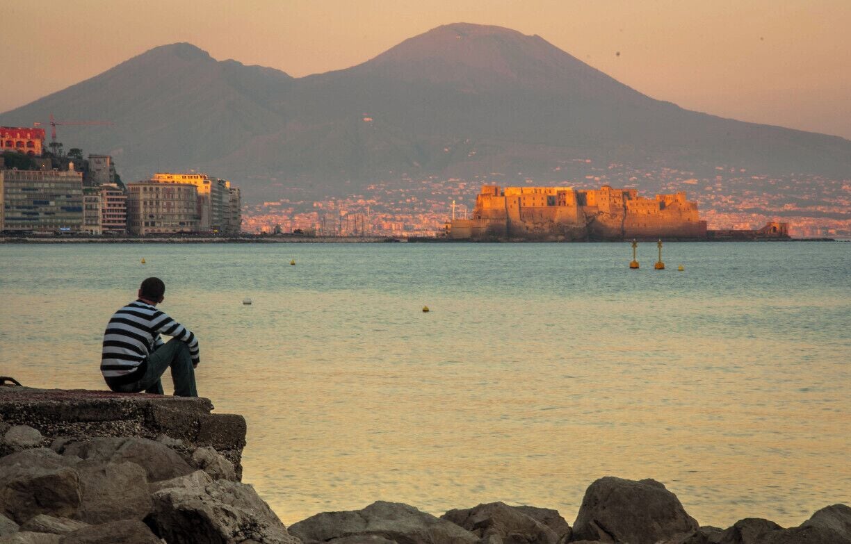 Um homem sentado à beira-mar, com uma montanha e um castelo ao fundo.