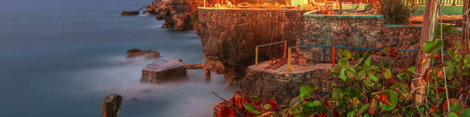Uma paisagem costeira com uma estrutura de pedra, um cais de madeira e um edifício iluminado.