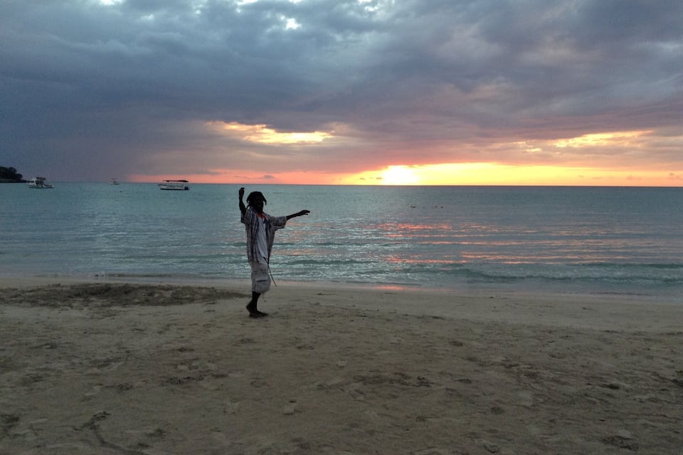Uma pessoa parada na praia, com o oceano e o pôr do sol ao fundo.