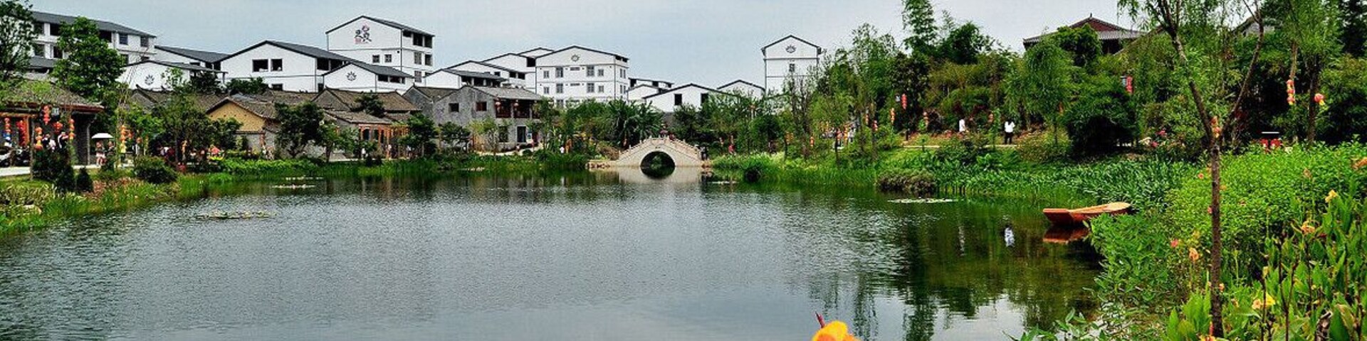 Um lago tranquilo com uma ponte de pedra, rodeado por vegetação exuberante e edifícios tradicionais.