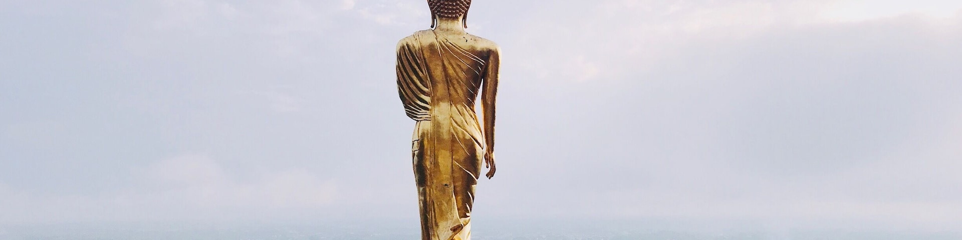 Uma grande estátua dourada de Buda sobre um pedestal, rodeada por uma estrutura de pedra com vista para o horizonte da cidade ao fundo.