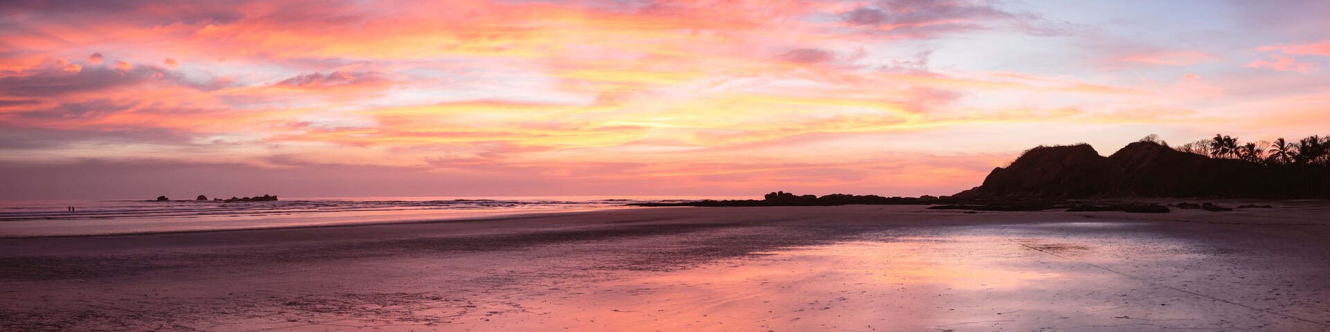 Uma praia ao pôr do sol com céu rosado e alaranjado, águas calmas e formações rochosas ao longe.