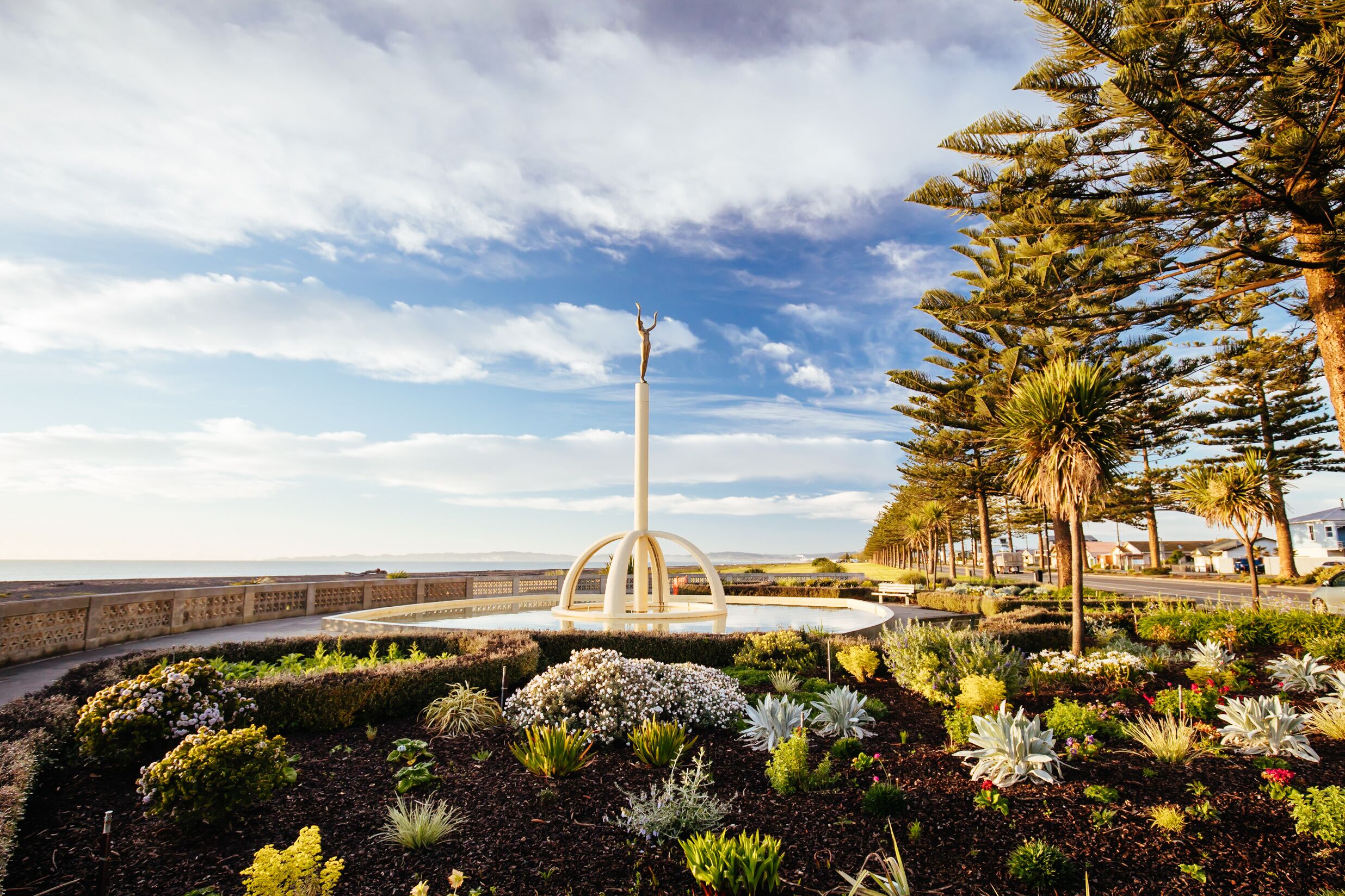 Um jardim paisagístico com uma escultura central, rodeado por plantas e árvores diversas, sob um céu parcialmente nublado.