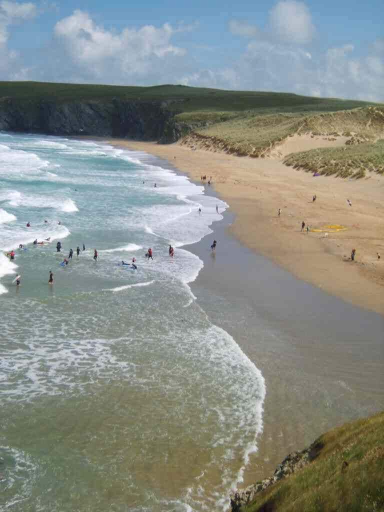 Uma praia costeira com ondas, pessoas e dunas gramadas.