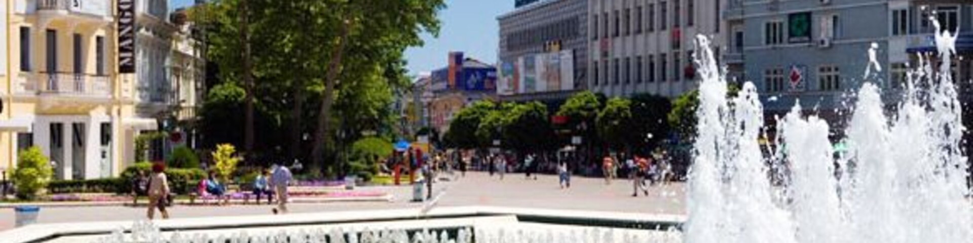 Uma fonte em uma praça, com edifícios e árvores ao redor.