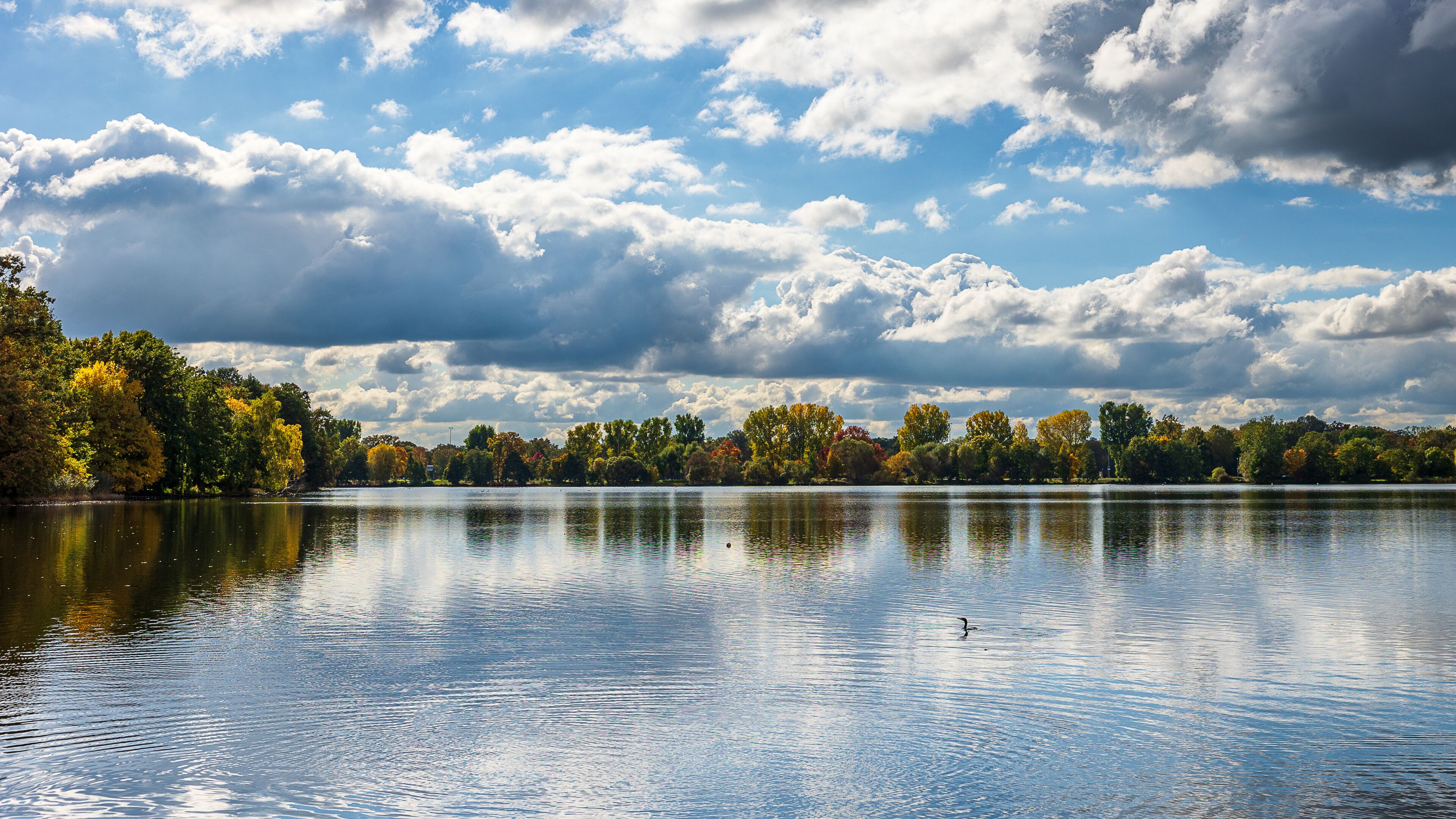 Um lago tranquilo com reflexos de árvores e nuvens, rodeado por uma floresta com folhagem outonal.