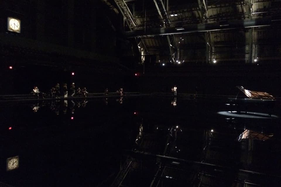 Um espaço interno com iluminação suave, um chão espelhado, um palco com algumas pessoas e uma grande estrutura industrial ao fundo.