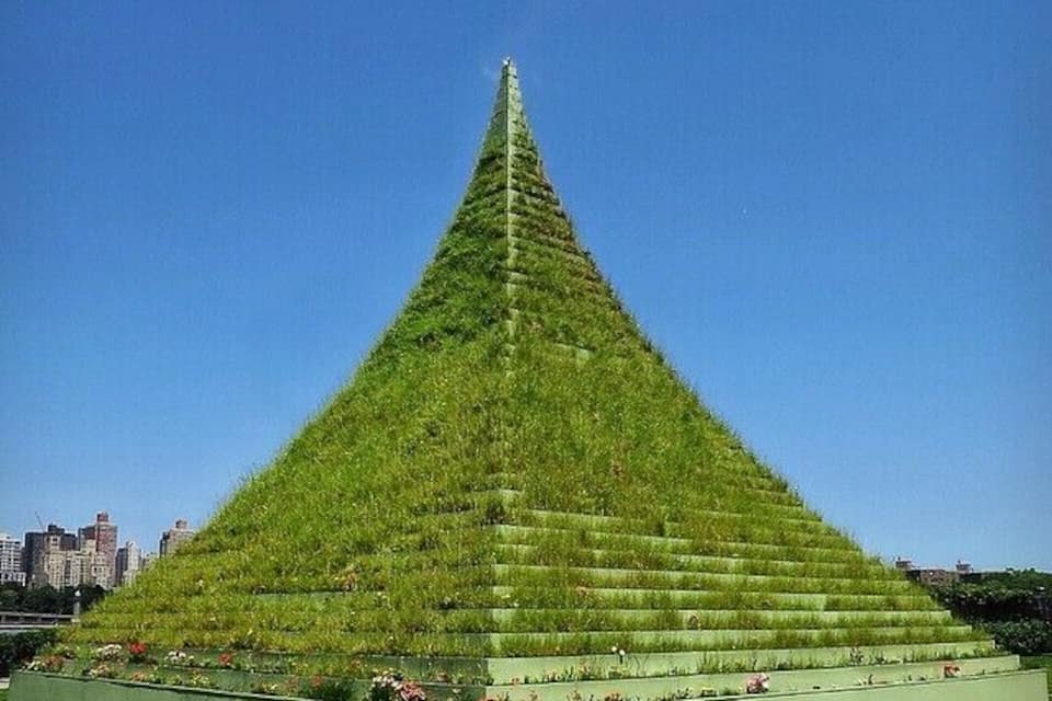 Uma grande estrutura em formato de pirâmide com superfície gramada, cercada por um muro baixo e um céu azul claro.