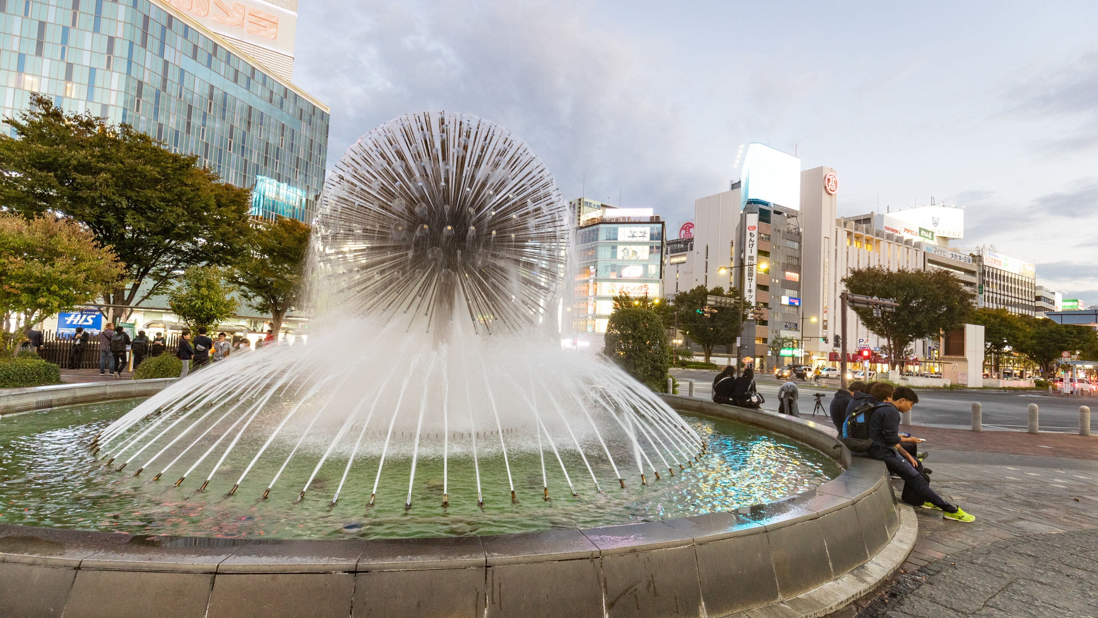 Uma fonte com jatos de água em praça pública, com edifícios ao fundo.