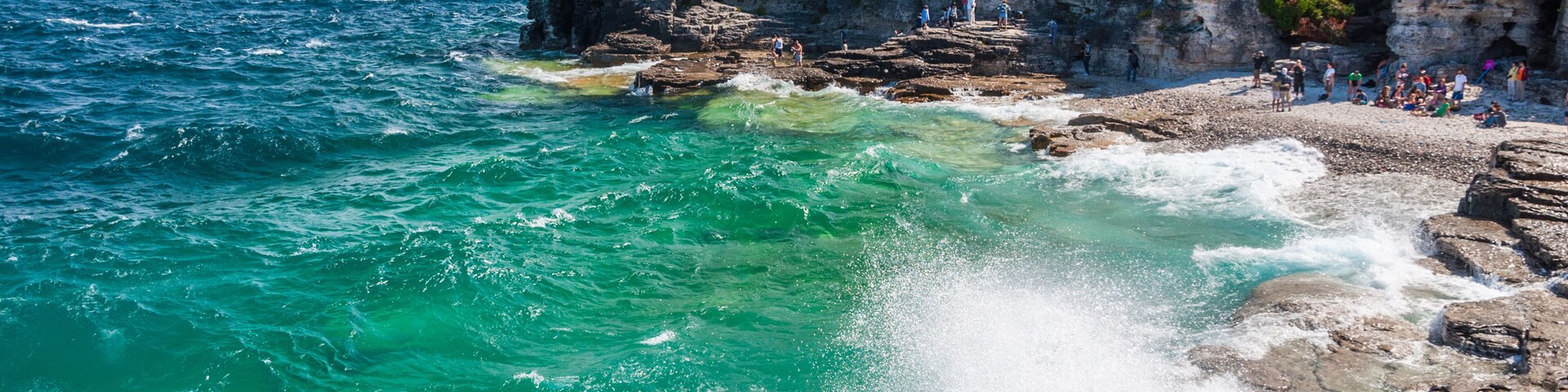 Litoral rochoso com águas turquesa cristalinas, um penhasco arborizado e pessoas aproveitando a praia.