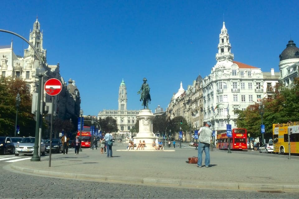 Um movimentado cruzamento urbano com edifícios históricos, uma estátua e ônibus de dois andares.