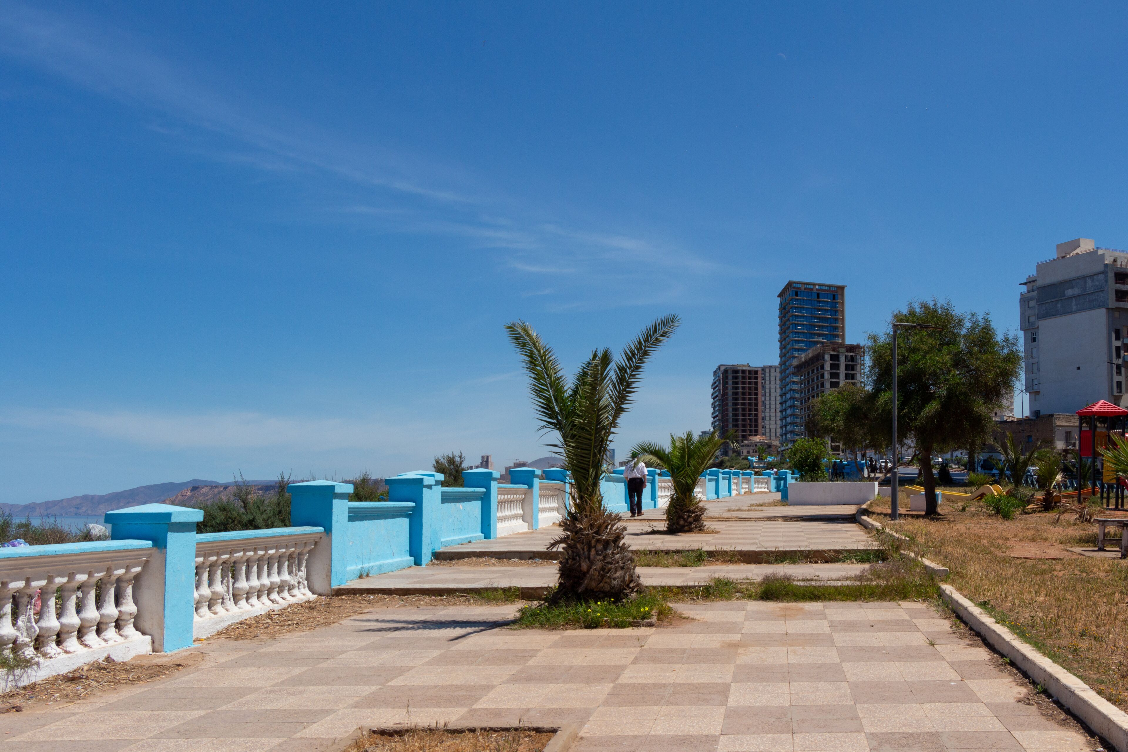 Uma passarela pavimentada com grades azuis, palmeiras e edifícios ao fundo.