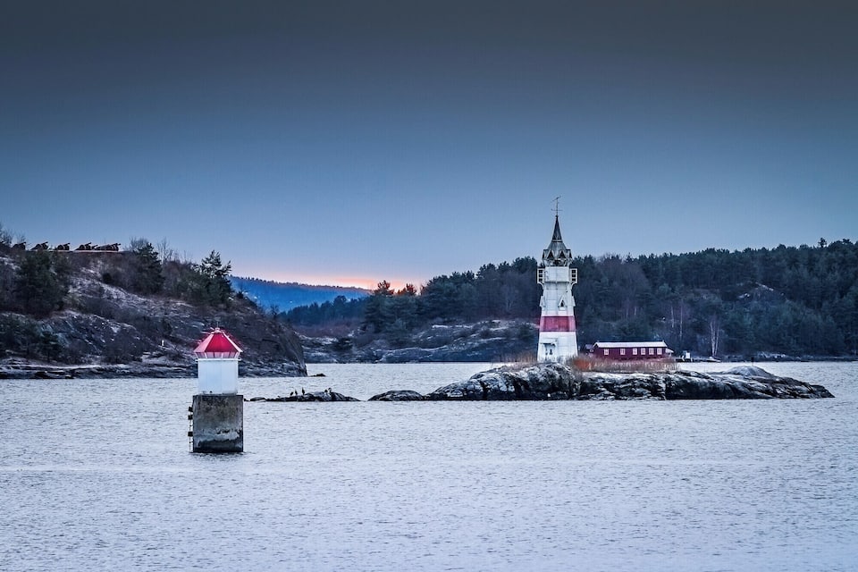 Um farol numa formação rochosa com o topo vermelho, rodeado por terreno coberto de neve e uma colina arborizada ao fundo.