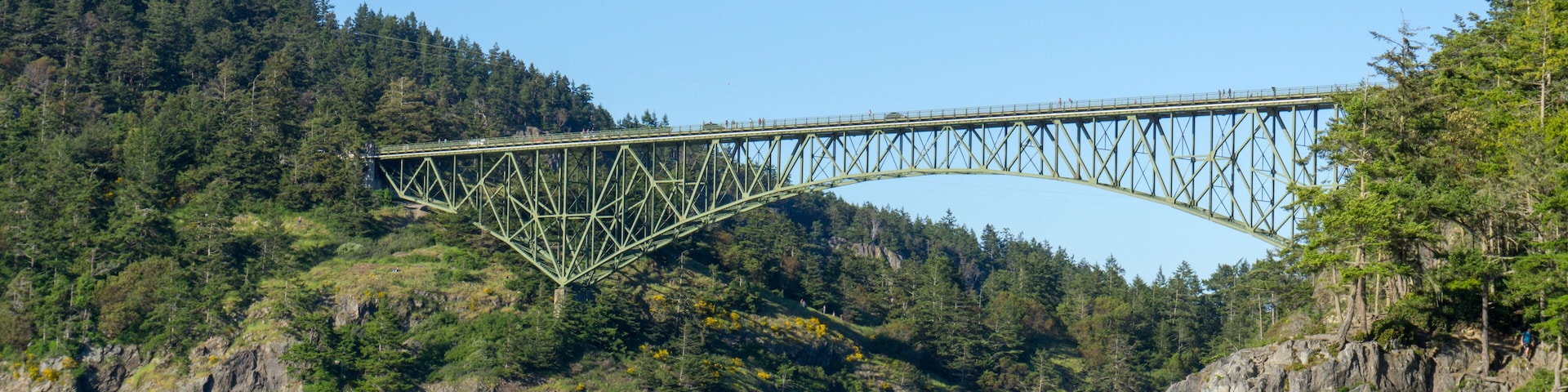 Uma ponte de aço sobre um rio, rodeada por colinas arborizadas e uma praia de pedras com pessoas.