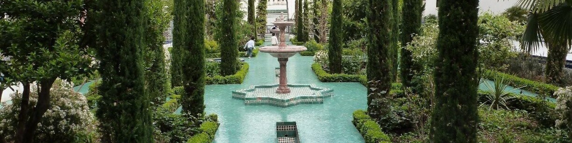 Um jardim formal com uma fonte central, caminhos simétricos e árvores altas.