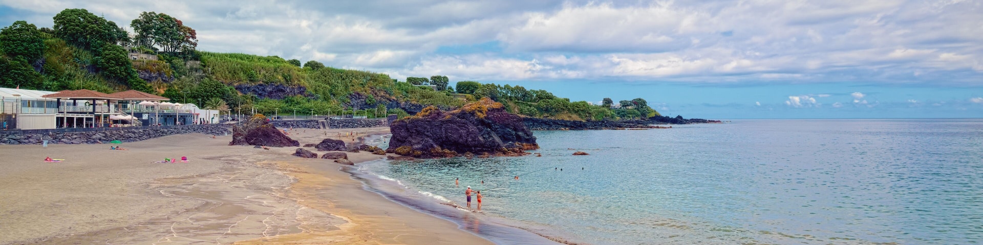 Uma praia com águas cristalinas, um afloramento rochoso e um edifício de fachada branca.