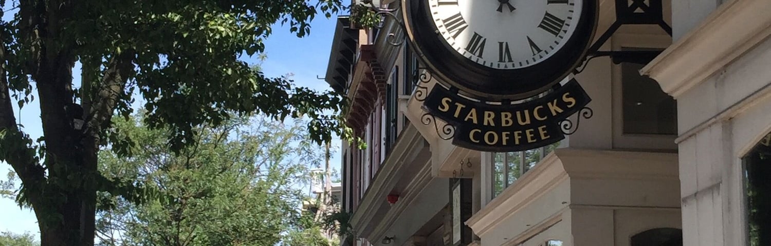 Cena de rua com um relógio marcando 12:05, uma placa da Starbucks Coffee e pessoas sentadas em mesas externas.