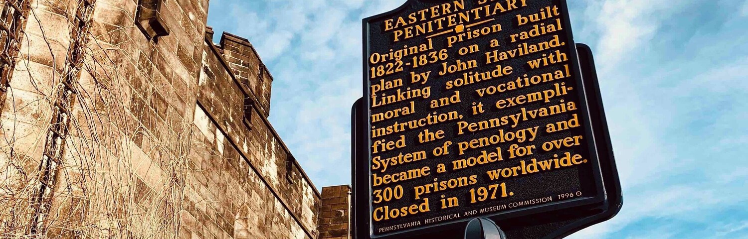 Uma placa histórica com informações sobre a Penitenciária Estadual do Leste, mencionando seu período de construção, fundadores e sua influência em sistemas prisionais mundiais.