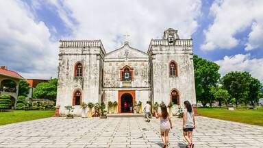 Igreja histórica com um amplo arco central e duas torres, rodeada por um pátio pavimentado e vegetação.