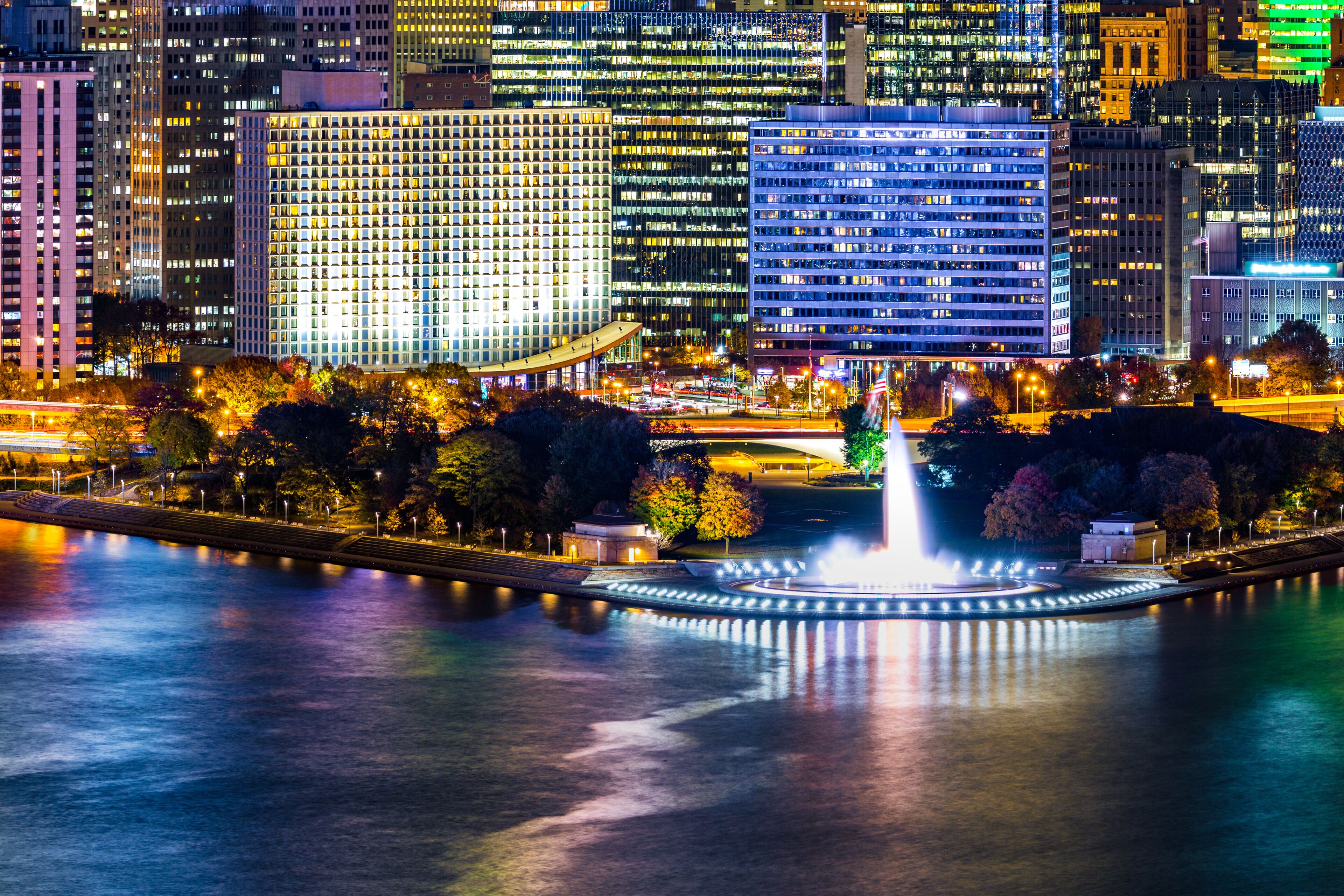Uma paisagem urbana noturna com edifícios iluminados, um rio e uma fonte.